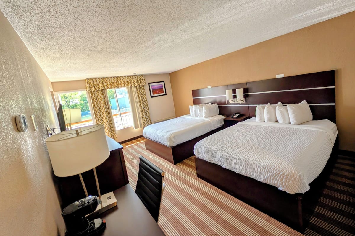 Quarto de hotel com duas camas, uma escrivaninha com abajur, telefone e janela com cortinas.