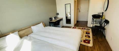 Quarto de hotel com uma cama grande, uma escrivaninha com cadeira, um espelho e uma kitchenette pequena.