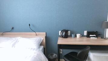 Quarto de hotel com cama, escrivaninha com cafeteira, lixeira e uma cadeira.