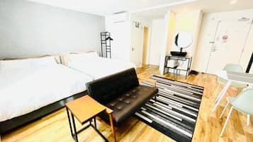 Quarto de hotel com cama, sofá de couro preto, mesa de madeira pequena, tapete listrado preto e branco, espelho redondo e pia branca.