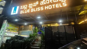 Exterior do Blue Bliss Hotels com um letreiro iluminado em destaque, uma escadaria que leva à entrada e uma loja de móveis por perto.