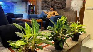 Um homem sentado em um sofá azul em um saguão com plantas em vasos e um ventilador.