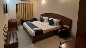 Quarto de hotel com cama, mesas de cabeceira, escrivaninha, cadeira, ventilador de teto e janela com cortinas.