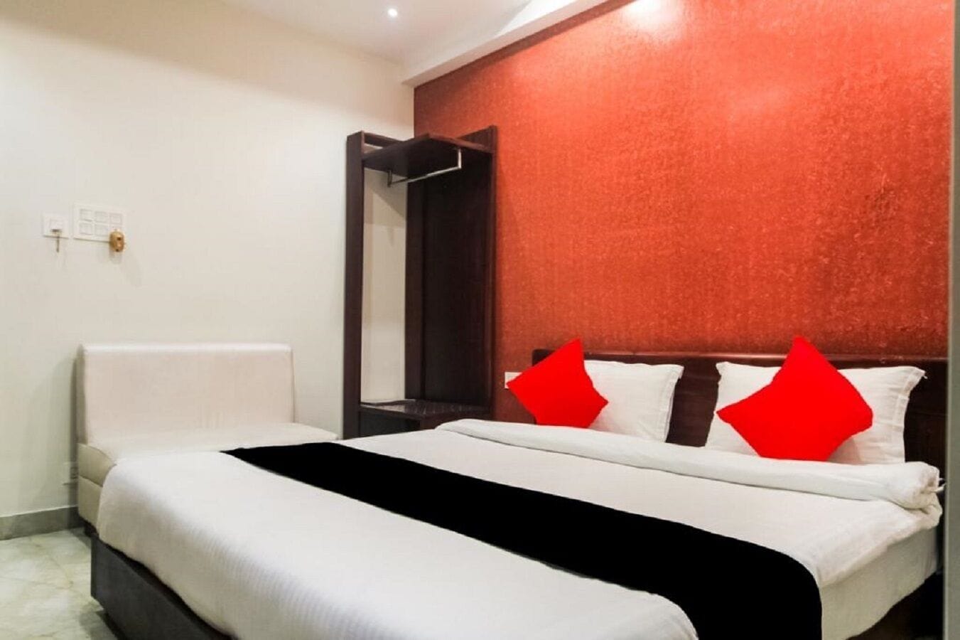 Quarto de hotel com cama, mesa de cabeceira, espelho e uma parede vermelha de destaque.
