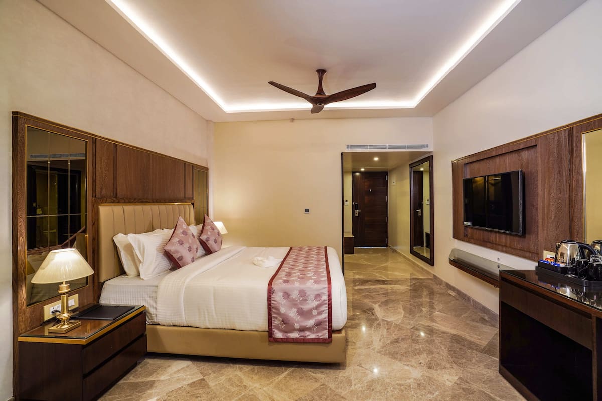 Quarto de hotel com uma cama grande, mesa de cabeceira com abajur, ventoinha no teto, TV na parede e espelho sobre a cama.