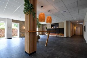 Um hall amplo com design moderno, destacando um pilar de madeira com vegetação, duas luminárias pendentes laranja e uma área de estar com cadeiras.