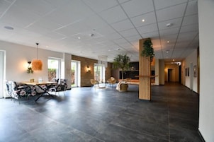 Um hall de hotel espaçoso e moderno, com pé direito alto, piso ladrilhado escuro e diferentes áreas de estar, incluindo uma mesa de jantar com cadeiras e um espaço lounge com sofás.