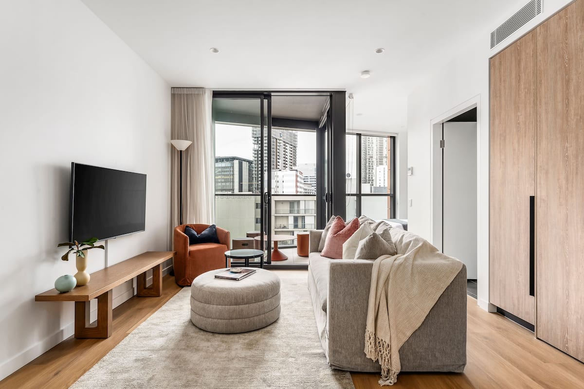 Uma sala de estar moderna com sofá, banquinho e banco, uma TV de tela plana e uma janela grande com vista para a cidade.