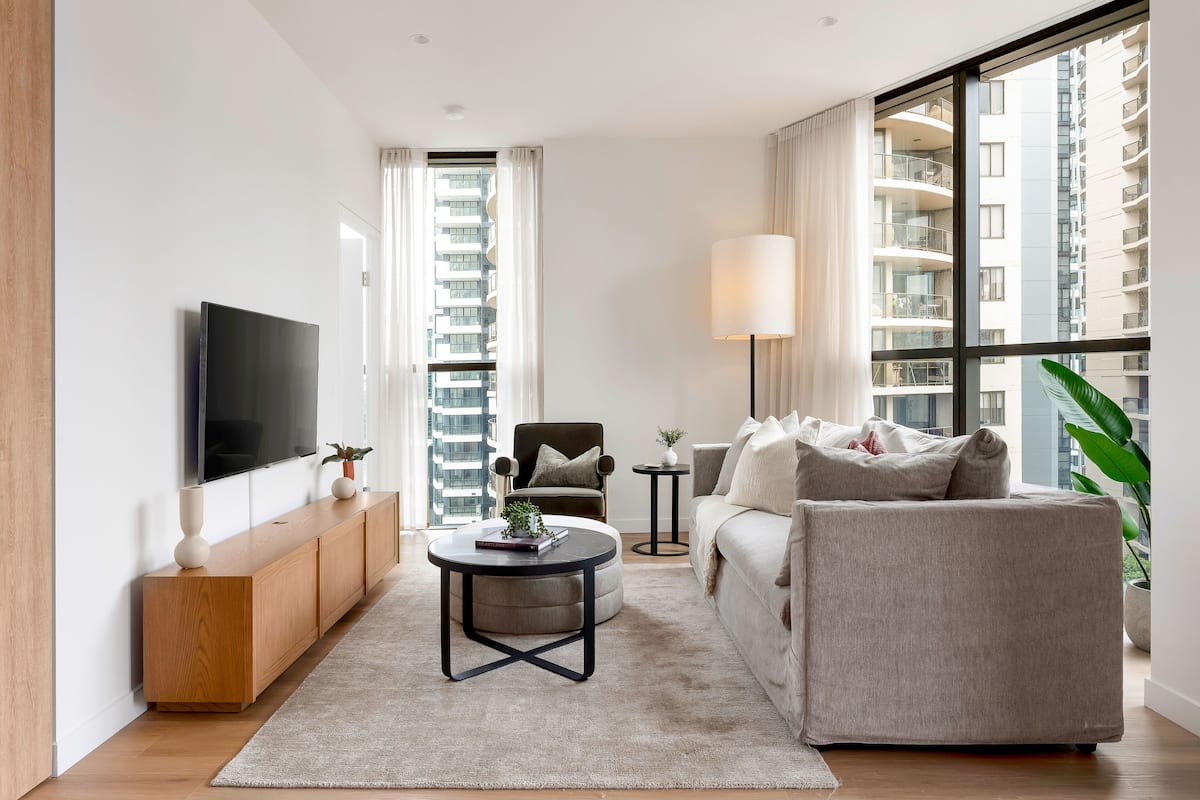 Uma sala de estar moderna com televisão de tela plana, um sofá, uma mesa de centro e vista para edifícios altos através de grandes janelas.