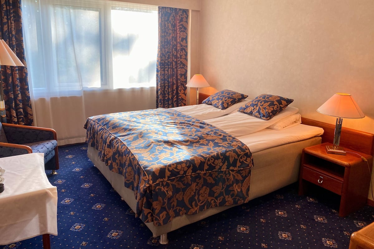 Quarto de hotel com uma cama grande, duas mesas de cabeceira, uma cadeira e uma janela com cortinas.