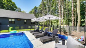 Piscina com cadeiras de descanso e um guarda-sol, cercada por uma vedação e árvores.