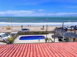 Vista da praia com piscina, cadeiras de praia, guarda-sóis e carros estacionados perto da orla.