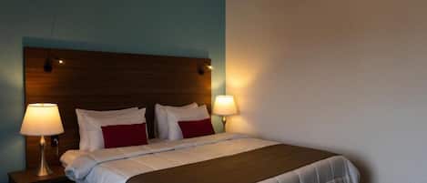 Cama bem arrumada com almofadas brancas e vermelhas, duas luminárias de cabeceira e cabeceira de madeira.