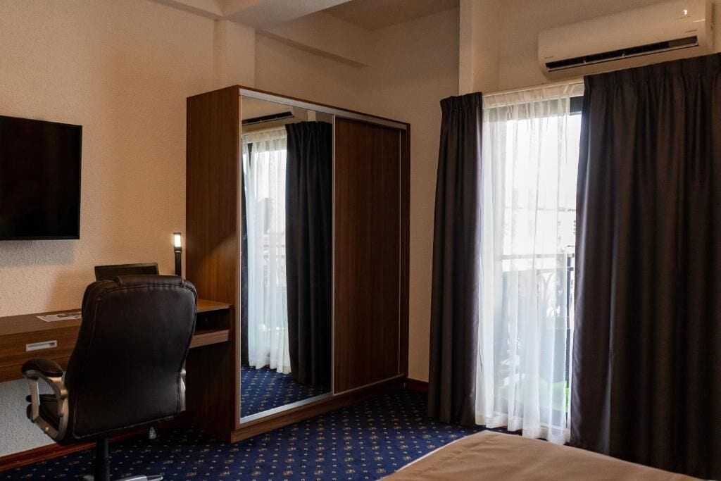 Quarto de hotel com cama, escrivaninha, cadeira, guarda-roupa, espelho e janela com cortinas.