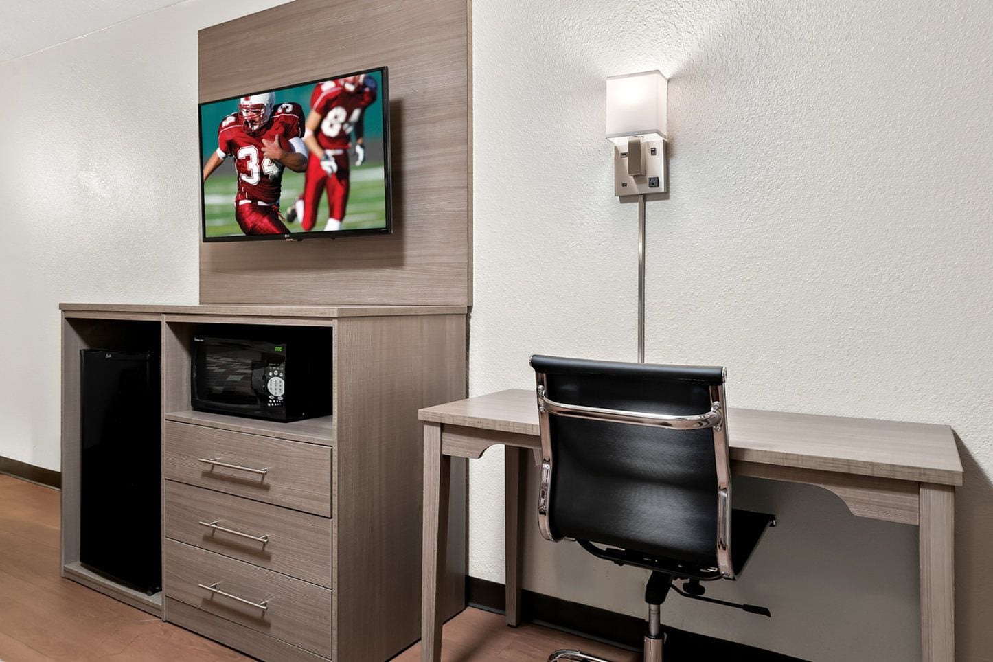 Quarto moderno com TV de tela plana fixada na parede, escrivaninha com cadeira e micro-ondas integrado.