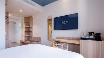 Quarto de hotel com cama, escrivaninha, cadeira, televisão e guarda-roupa.