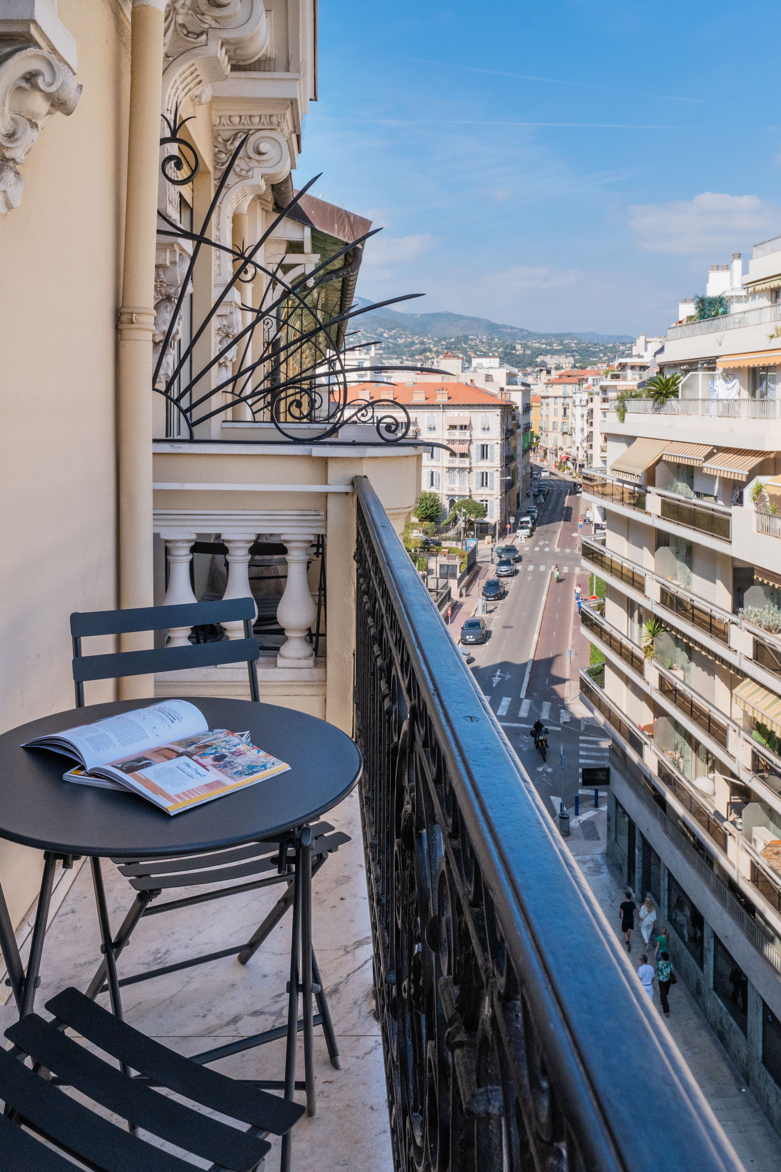 Varanda com mesa e cadeiras, com vista para uma rua com edifícios e uma montanha ao longe.