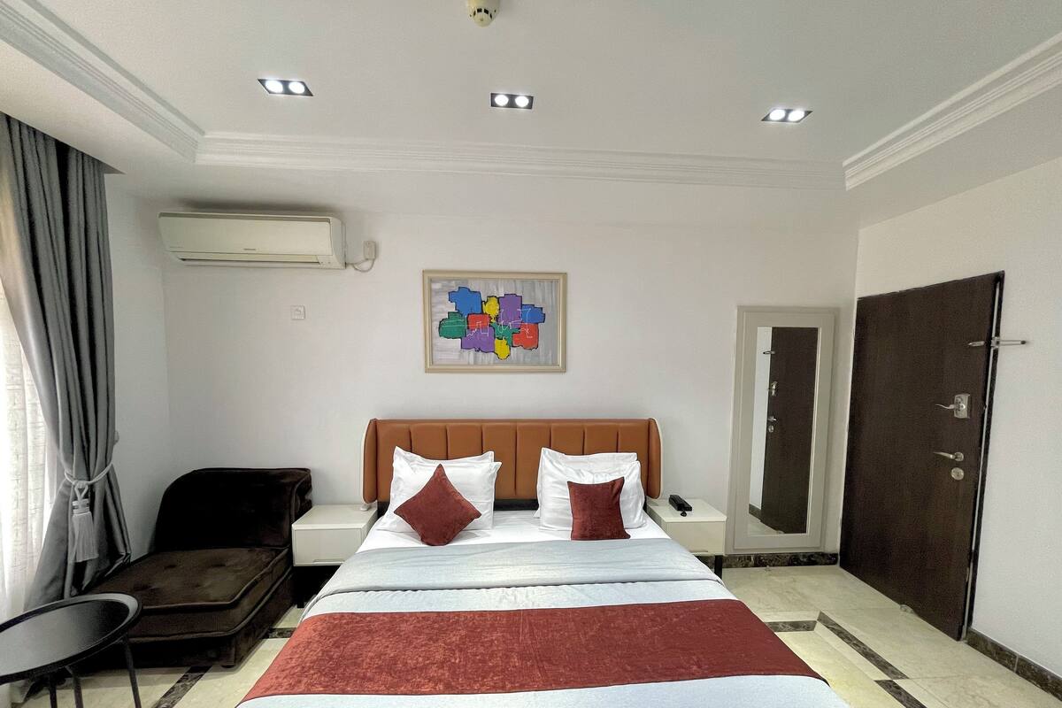 Quarto de hotel com cama grande, mesa de cabeceira, escrivaninha, cadeira, ar condicionado, porta e obra de arte na parede.