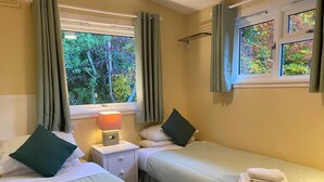 Quarto de hotel com duas camas, mesa de cabeceira com abajur e janela com cortinas.