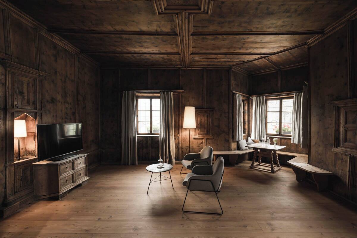 Um quarto rústico com paredes e piso de madeira, uma televisão, uma mesa com cadeiras e duas janelas com cortinas.