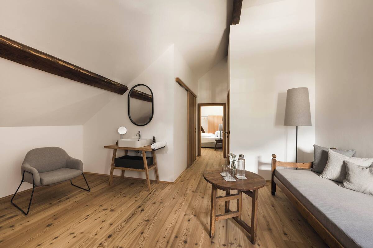 Quarto com piso de madeira, sofá, mesa pequena, cadeira, mesa de cabeceira, abajur e espelho.