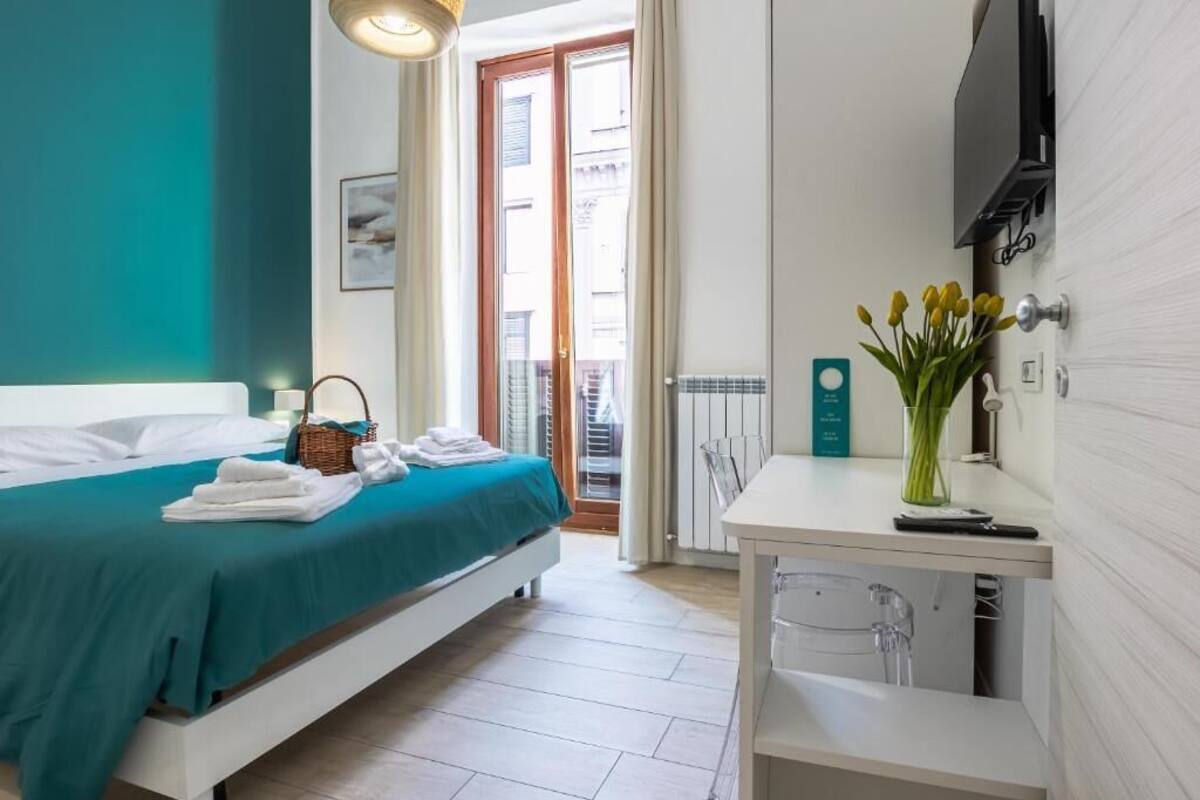 Quarto com cama, mesa de cabeceira com um vaso de flores e vista para o exterior.