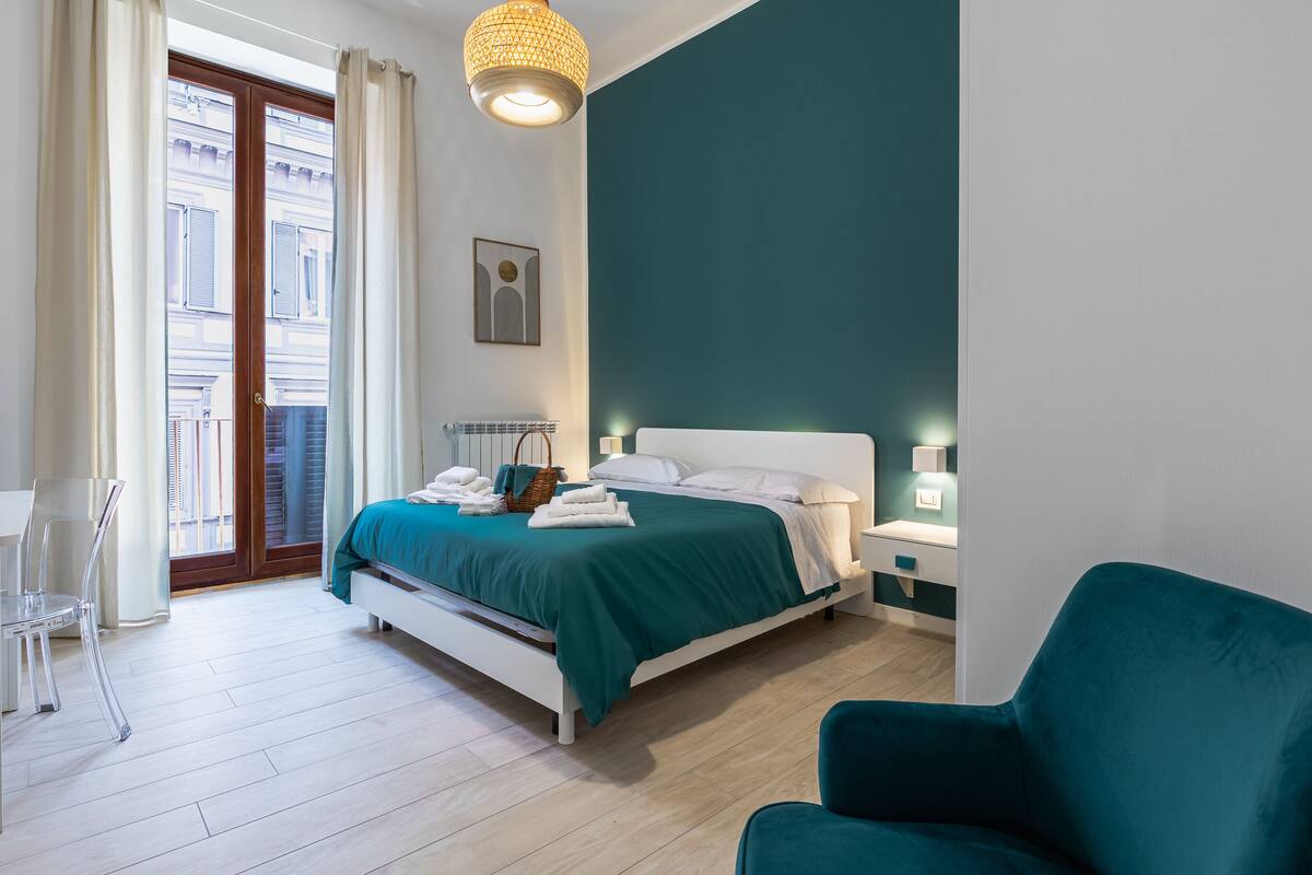 Quarto moderno com cama, mesa de cabeceira e uma cadeira. Há uma janela com cortinas, vista de prédios lá fora e um lustre.
