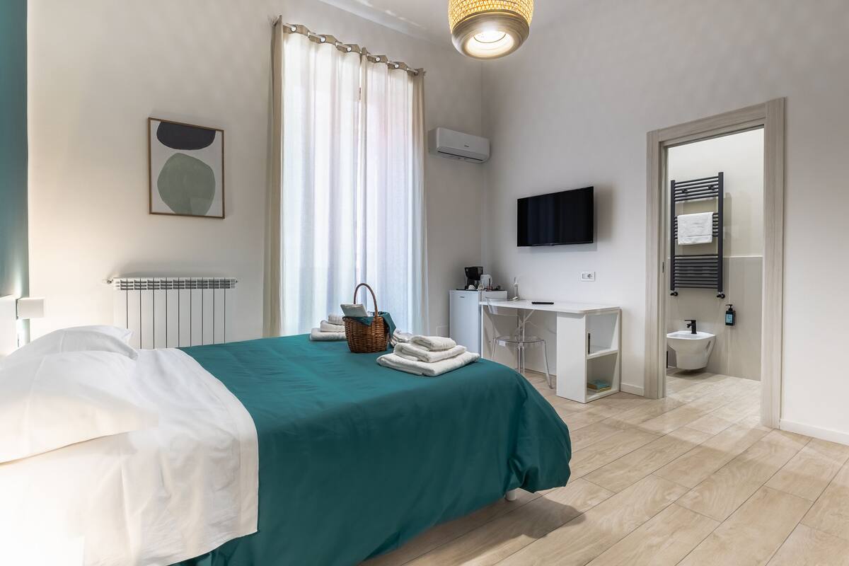 Quarto moderno com cama, escrivaninha, televisão e banheiro visível por uma porta aberta.