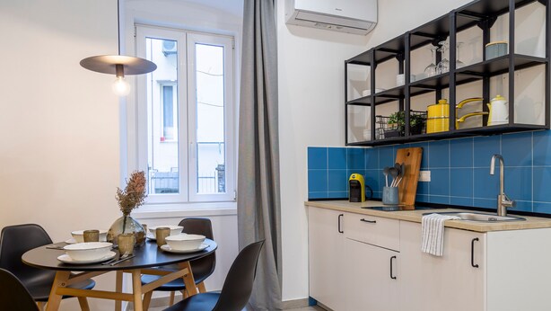 Uma cozinha moderna com área de jantar, com uma mesa redonda e cadeiras, uma estante embutida com utensílios de cozinha e uma janela com cortinas.