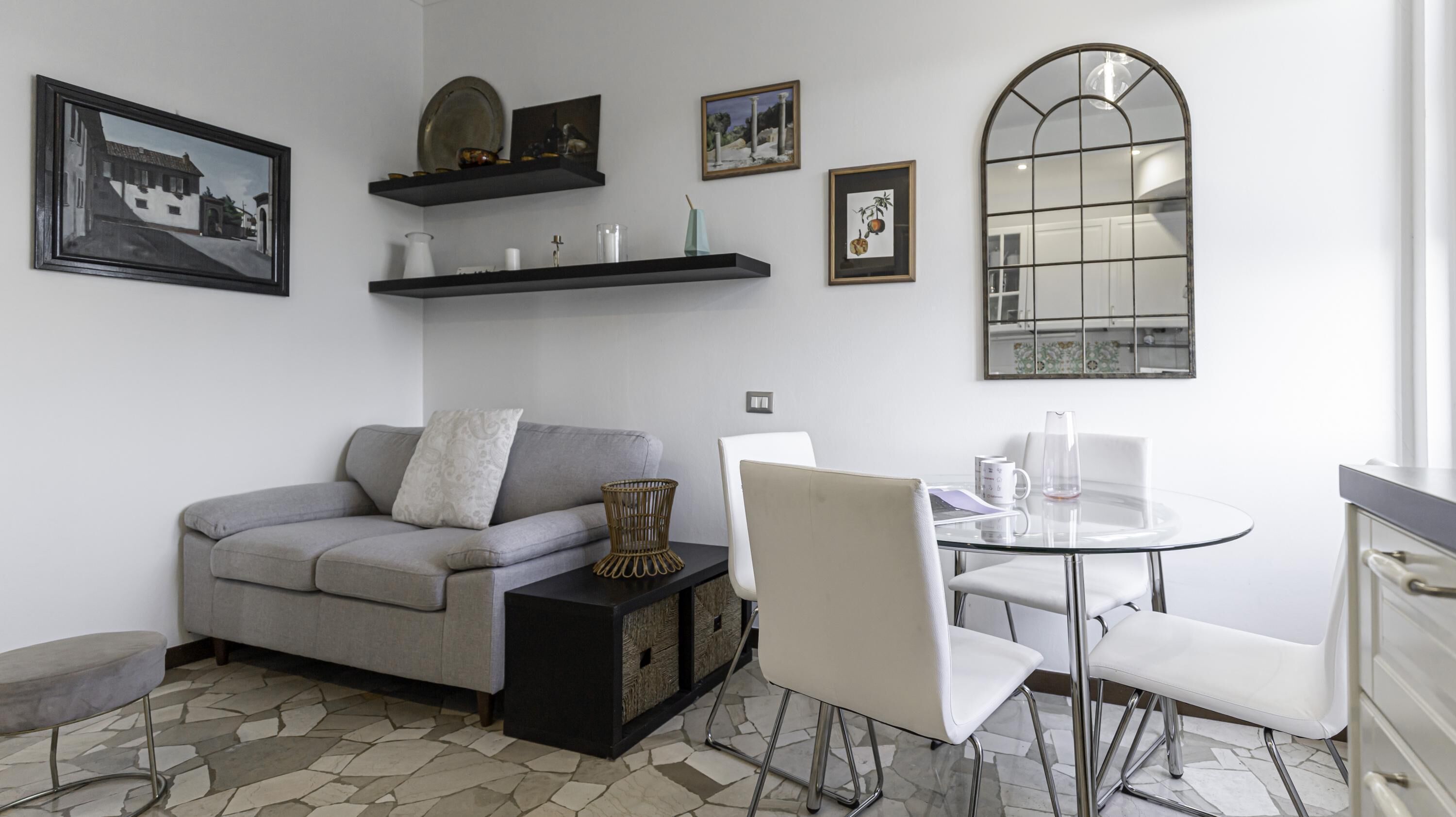 Uma sala de estar moderna com um sofá cinzento, cadeiras brancas, uma mesa de vidro e obras de arte nas paredes.