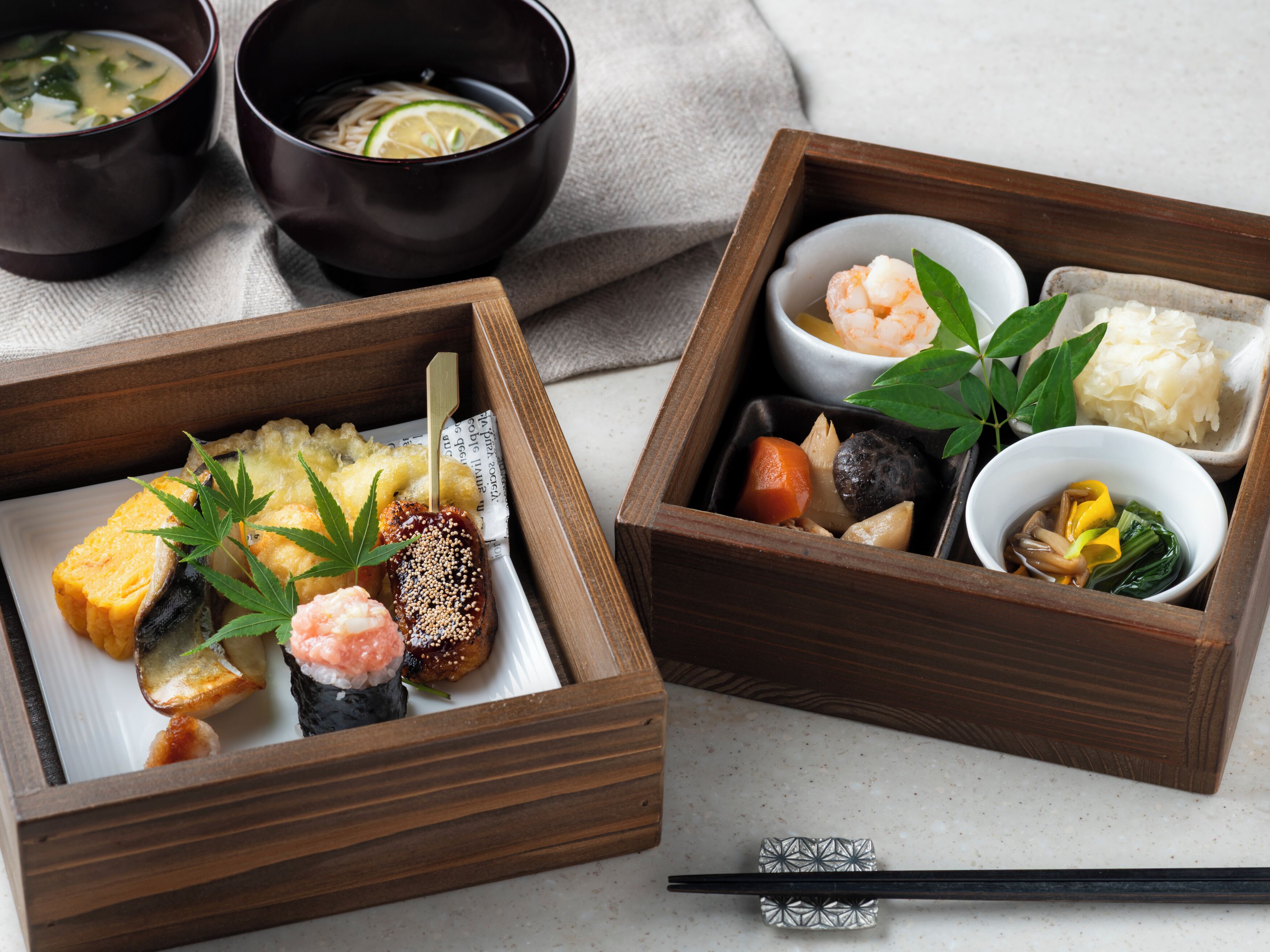 Duas caixas de madeira contendo diversos pratos japoneses, incluindo sushi e legumes, sobre uma mesa com um par de pauzinhos e duas tigelas de sopa.