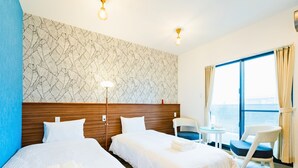 Quarto de hotel com duas camas, cabeceira de madeira, parede estampada, uma mesinha, uma cadeira e uma janela grande com cortinas.