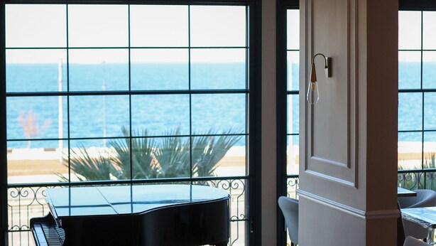 Quarto com piano, mesa e vista para o mar através de amplas janelas.