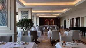 Uma sala de jantar formal com mesas preparadas para o serviço, um piano e uma grande árvore no centro.