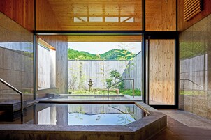 Um interior moderno com uma banheira de hidromassagem em vidro, teto de madeira e vista para um jardim com cerca de madeira e vegetação.