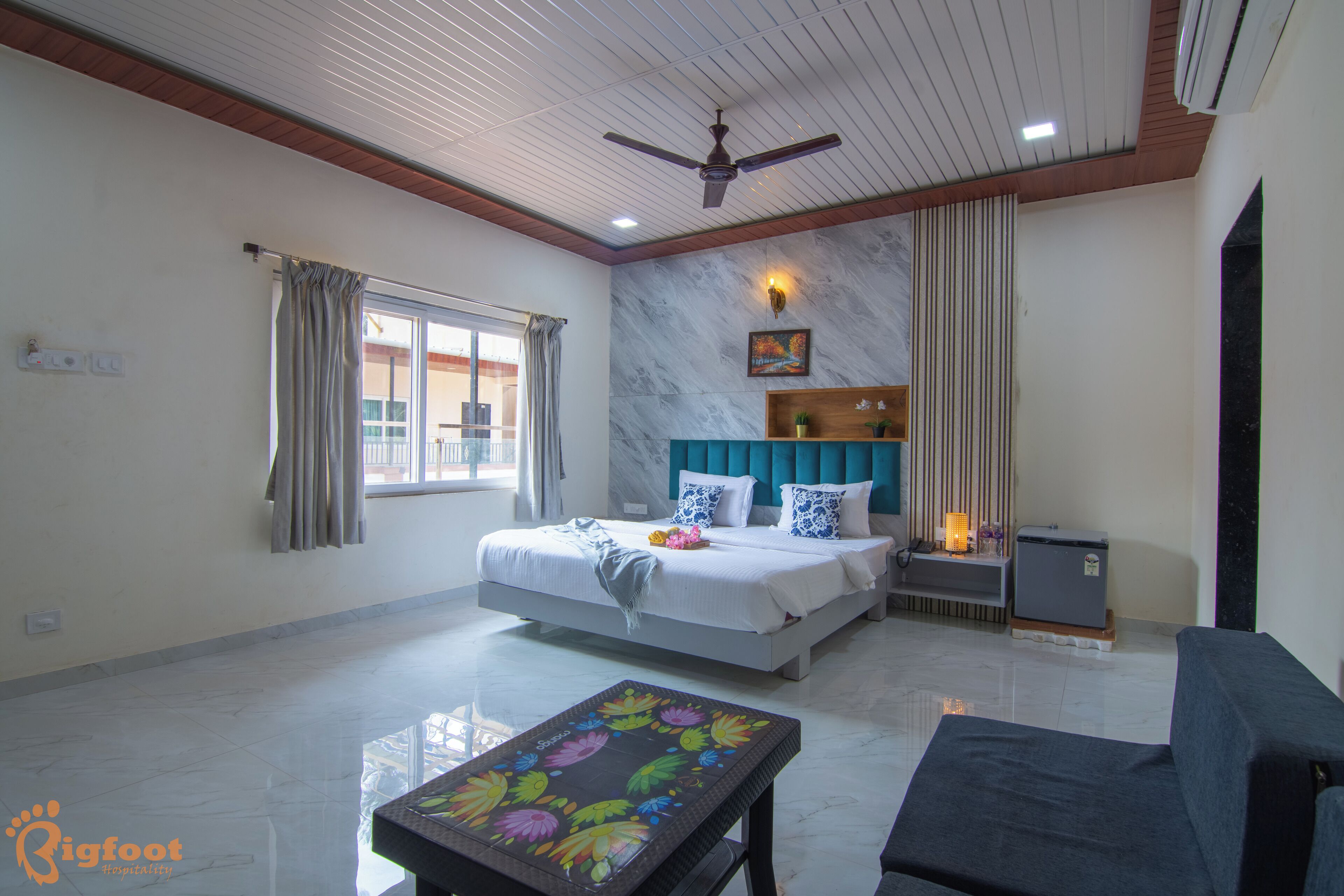 Quarto de hotel com cama, mesa pequena, sofá, ventoinha no teto e mesa de cabeceira com abajur.