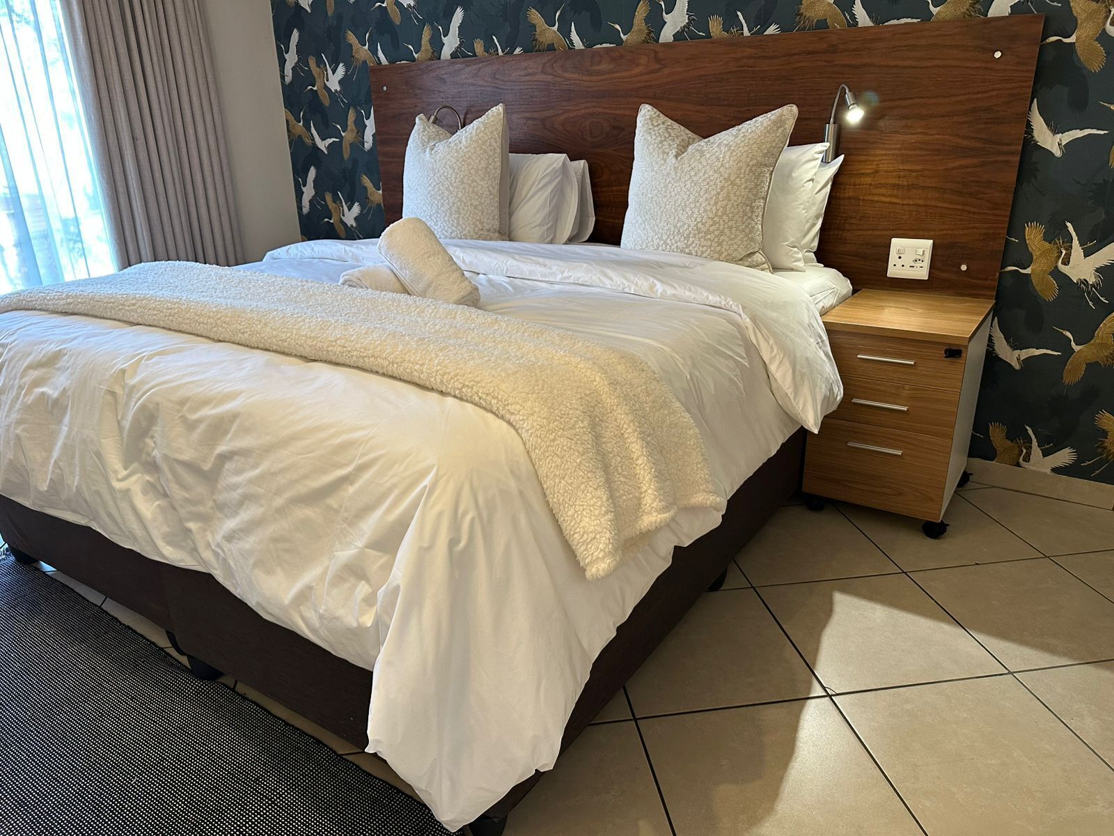 Quarto de hotel com uma cama grande, uma cabeceira de madeira, uma mesinha de cabeceira com gavetas e uma parede com padrão de pássaros.