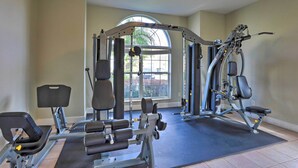 Uma academia com equipamentos de ginástica, um ventilador de teto e uma grande janela com vista para árvores.