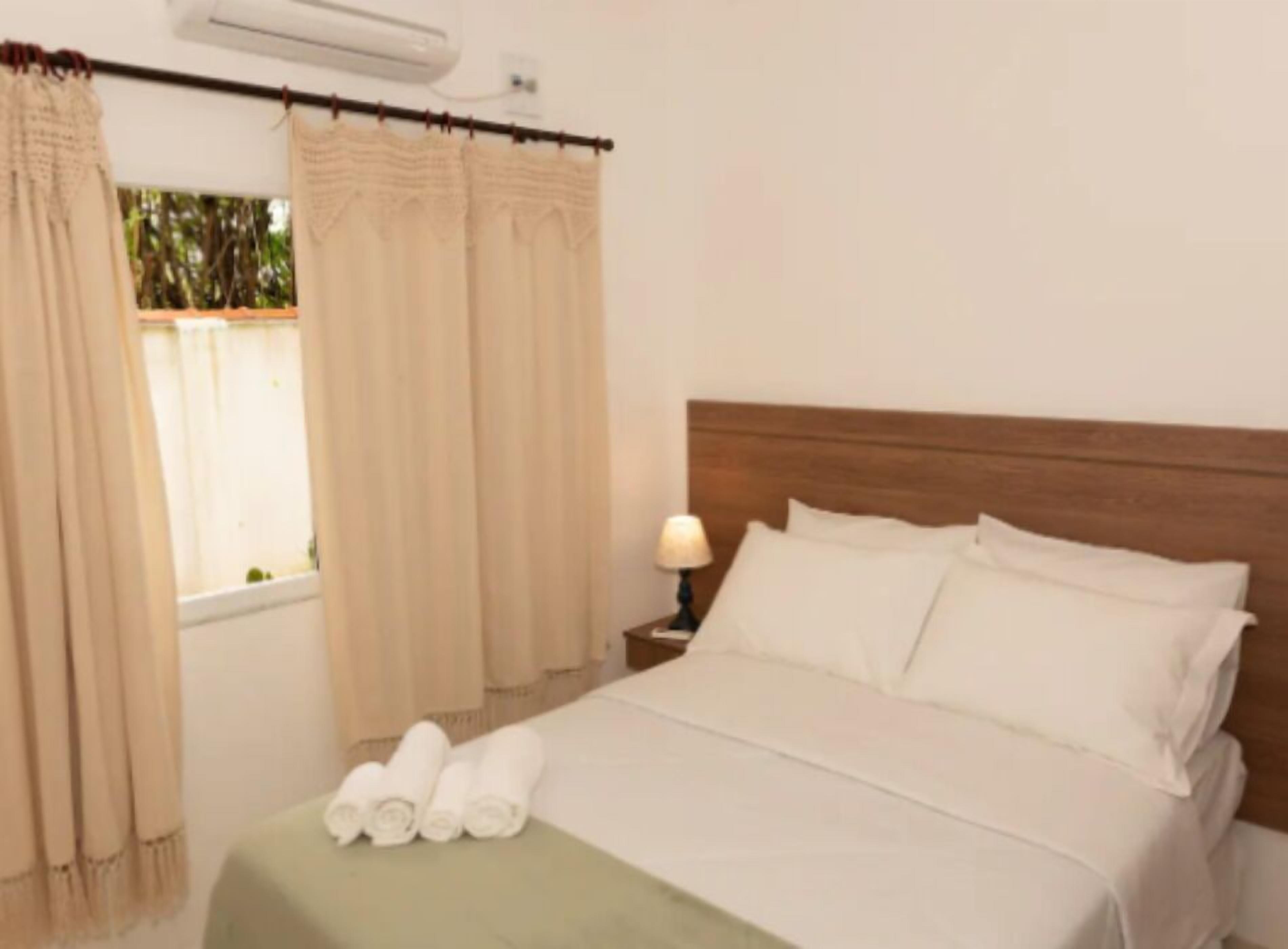 Cama bem arrumada com lençóis e almofadas brancos, cabeceira de madeira e mesa de cabeceira com abajur.