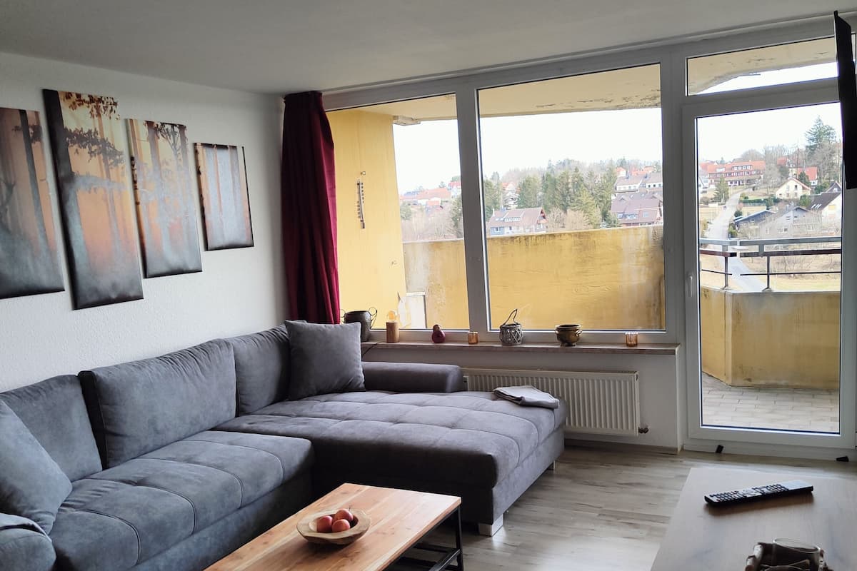 Uma sala de estar moderna com um sofá seccional cinza, uma mesa de centro de madeira e uma grande janela com vista para uma área residencial.
