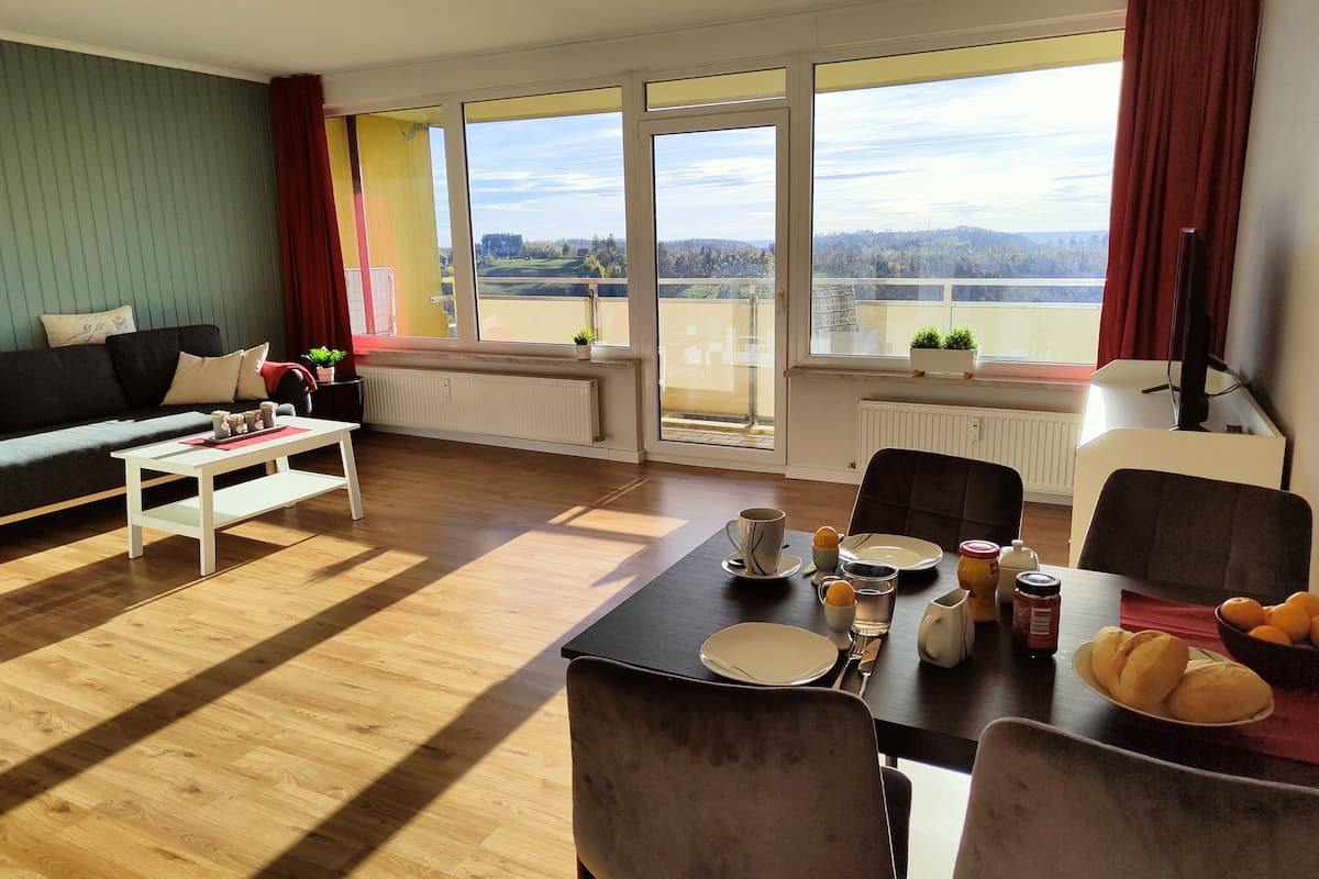 Uma sala de estar moderna com área de jantar, grandes janelas com vista para uma paisagem distante, um sofá, uma mesa de centro e uma mesa de jantar posta para refeição.