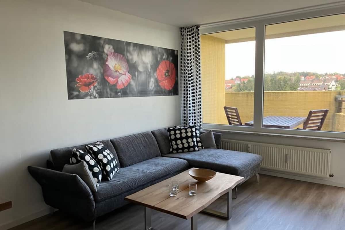 Uma sala de estar moderna com um sofá seccional cinza, uma mesa de centro de madeira e uma grande janela com vista para um horizonte urbano.