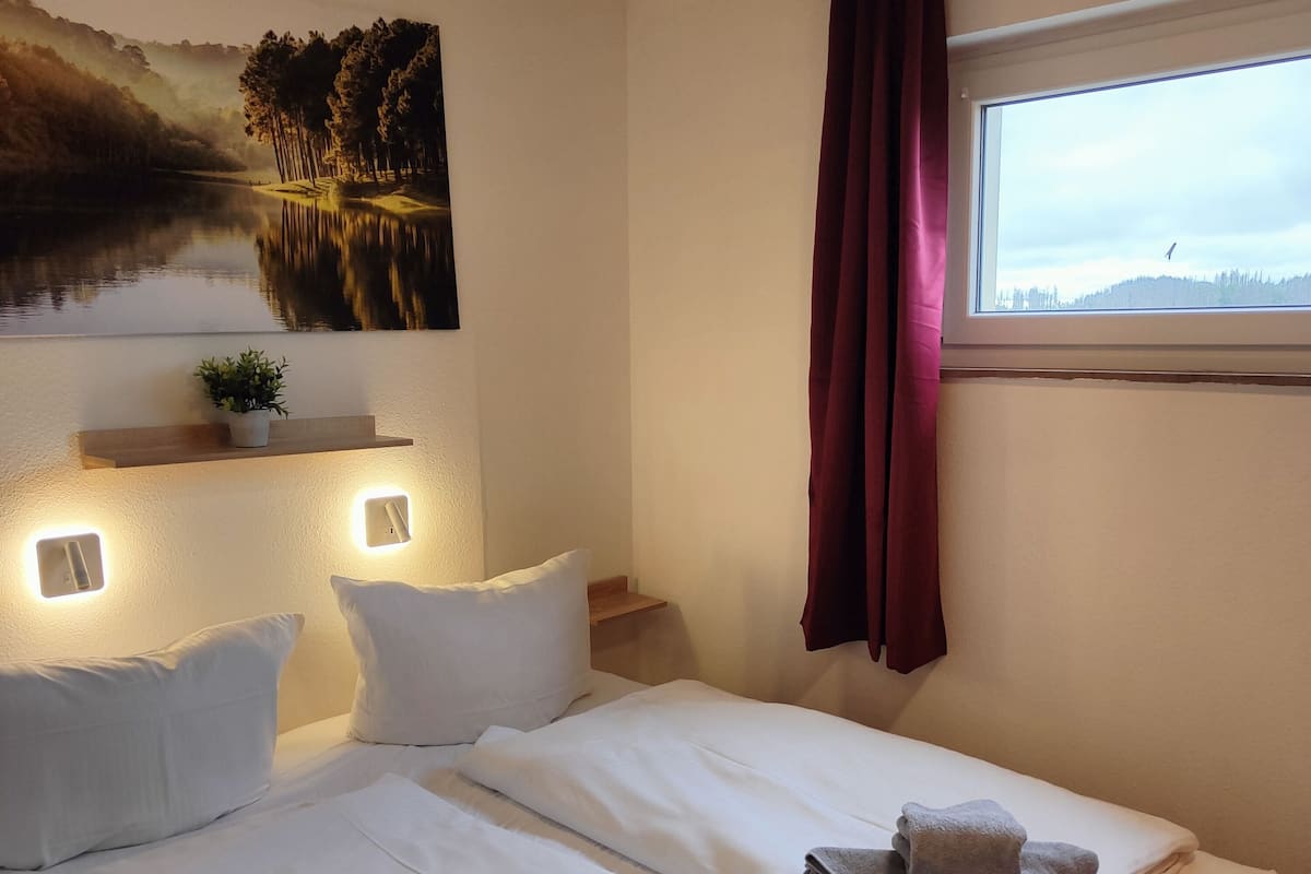 Quarto de hotel com duas camas, um quadro de uma floresta, uma janela com cortinas e vista para o céu.