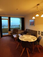 Uma sala de estar acolhedora com uma mesa redonda, duas poltronas, um sofá e vista para a cidade através de amplas janelas.