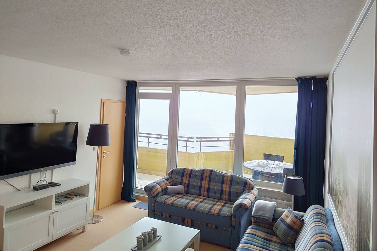 Uma sala de estar com televisão de tela plana, um sofá, uma mesa de centro e uma varanda com mesa e cadeiras.