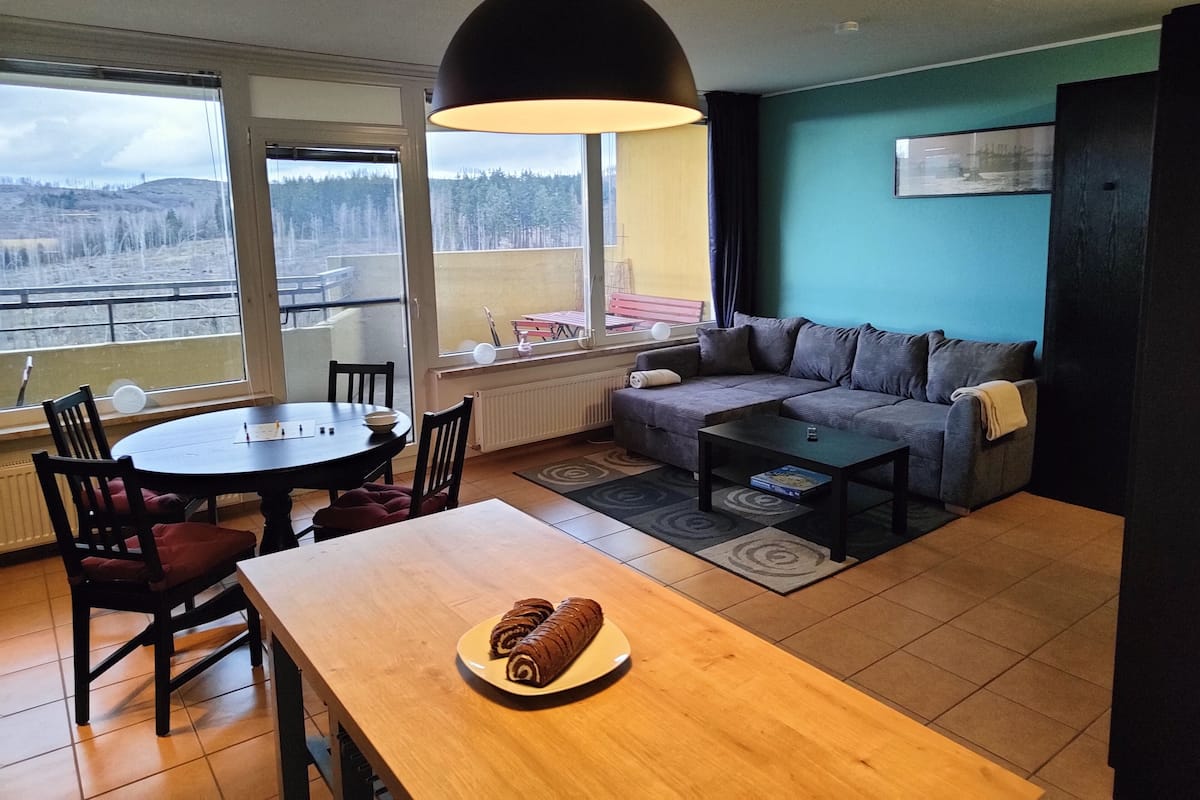 Uma sala de estar com sofá, uma mesa de jantar com cadeiras, uma mesa de centro e uma janela grande com vista para árvores e edifícios.