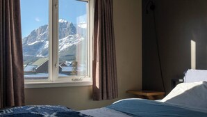 Quarto com cama, janela com cortinas, mesa de cabeceira e vista para montanhas nevadas.