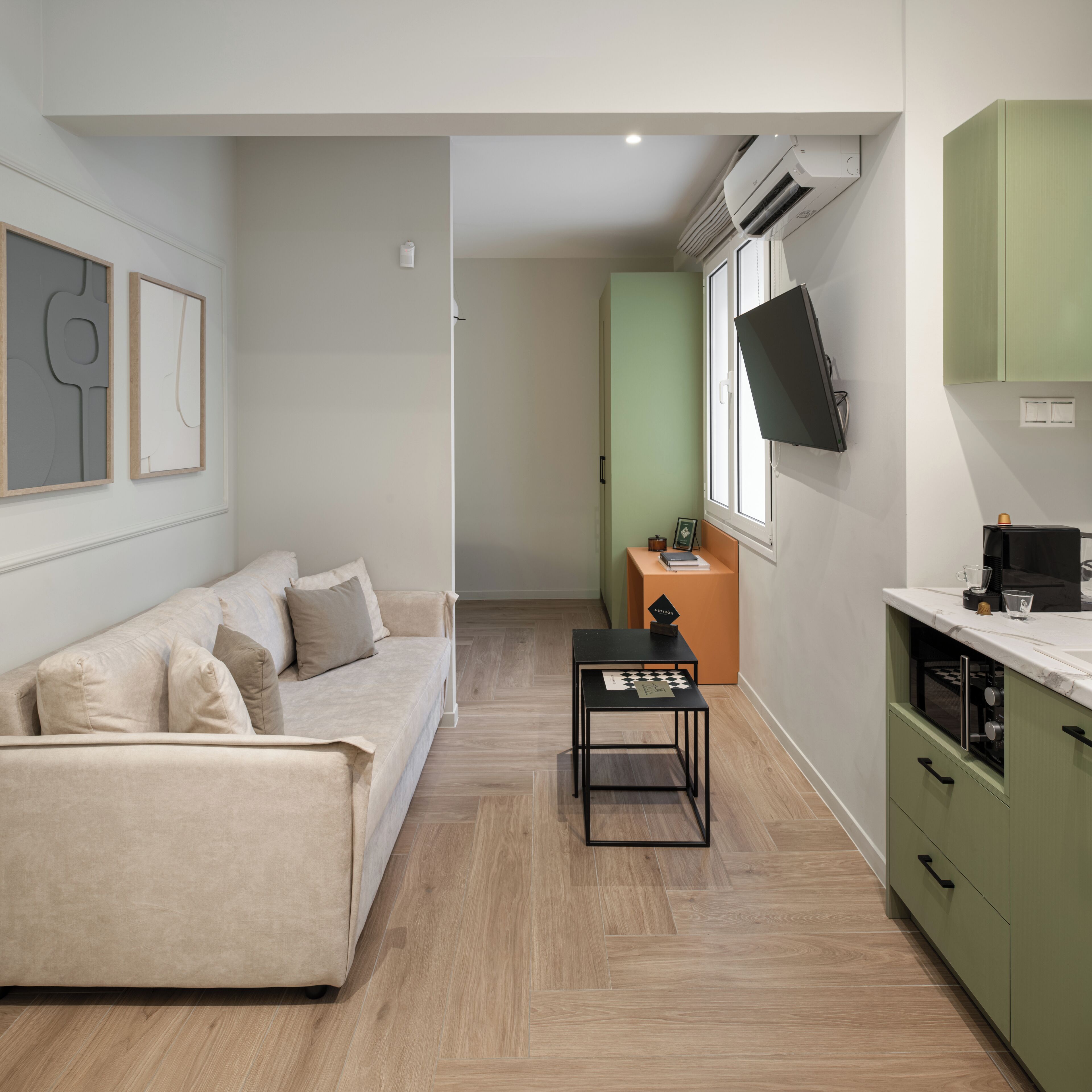 Um espaço de estar compacto com sofá, uma mesinha, área de cozinha com armários e uma TV fixada na parede.