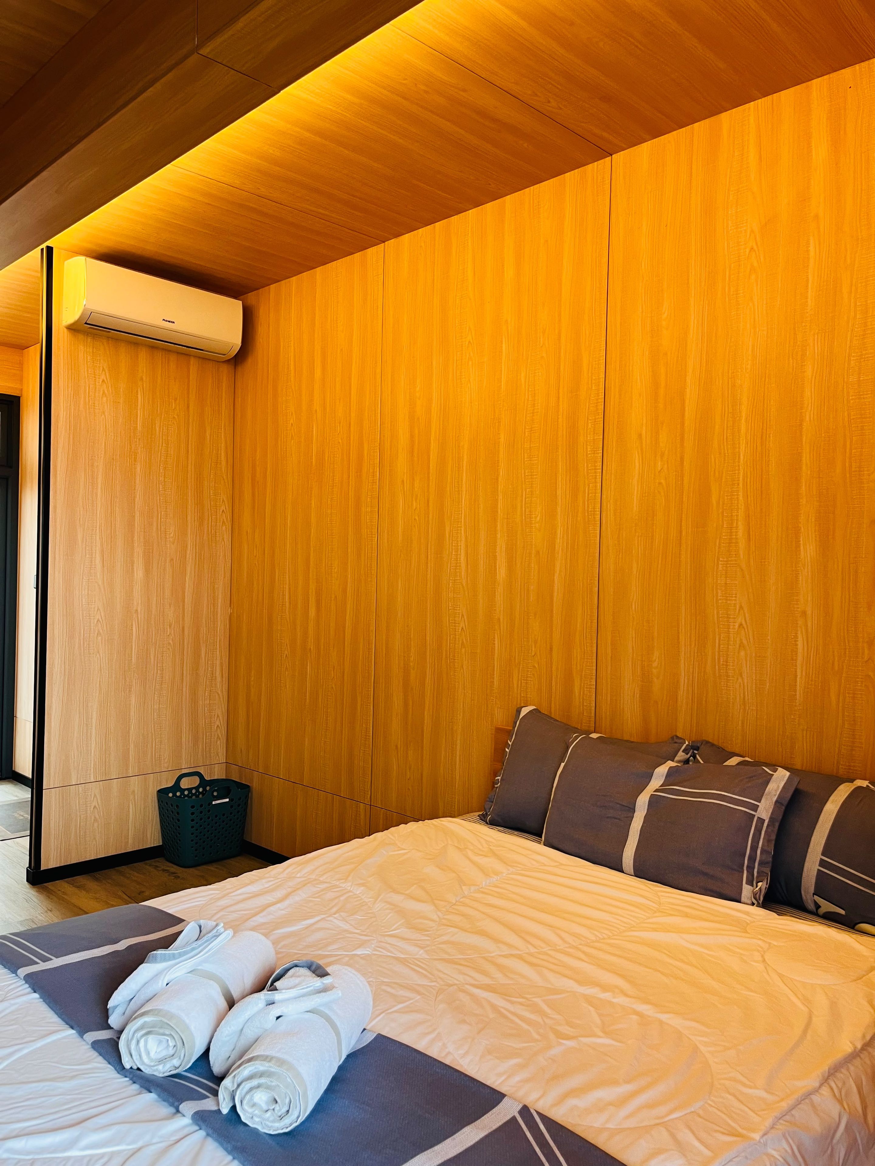 Quarto de hotel com parede de madeira, cama com colcha branca e cinza, duas almofadas, um cesto de cor escura e um aparelho de ar condicionado.
