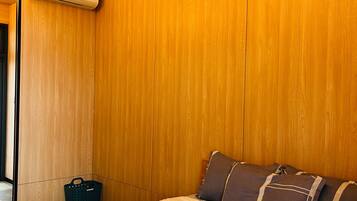 Quarto de hotel com parede de madeira, cama com colcha branca e cinza, duas almofadas, um cesto de cor escura e um aparelho de ar condicionado.